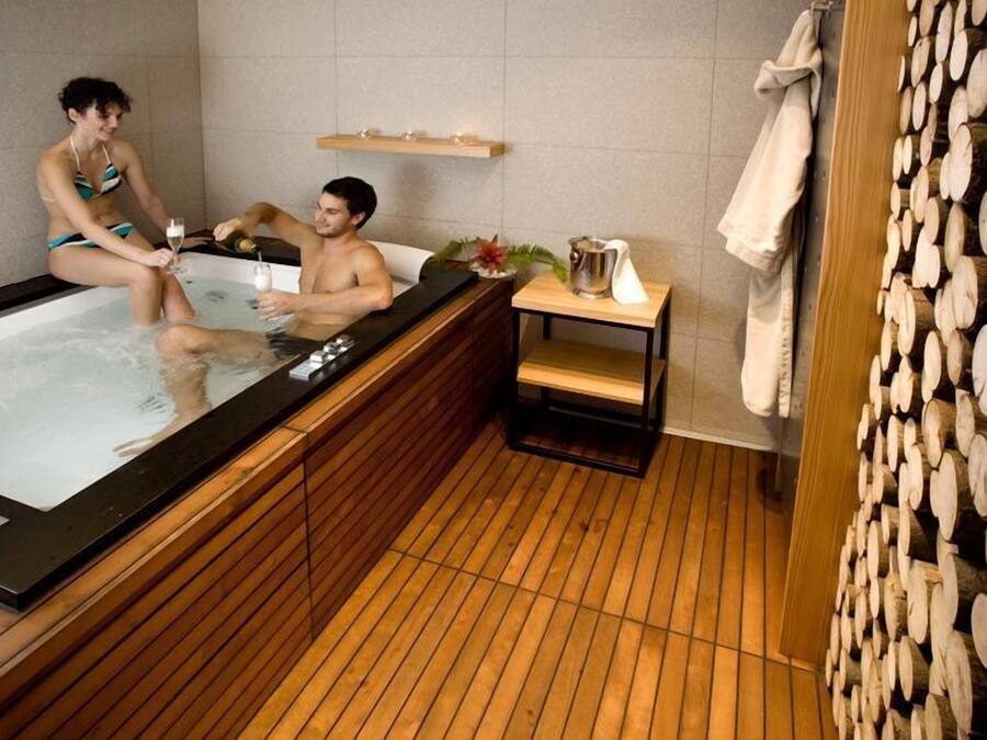 Quarto com piso de madeira, banheira de hidromassagem, uma pequena mesa com uma garrafa e uma planta, e um porta-toalhas.
