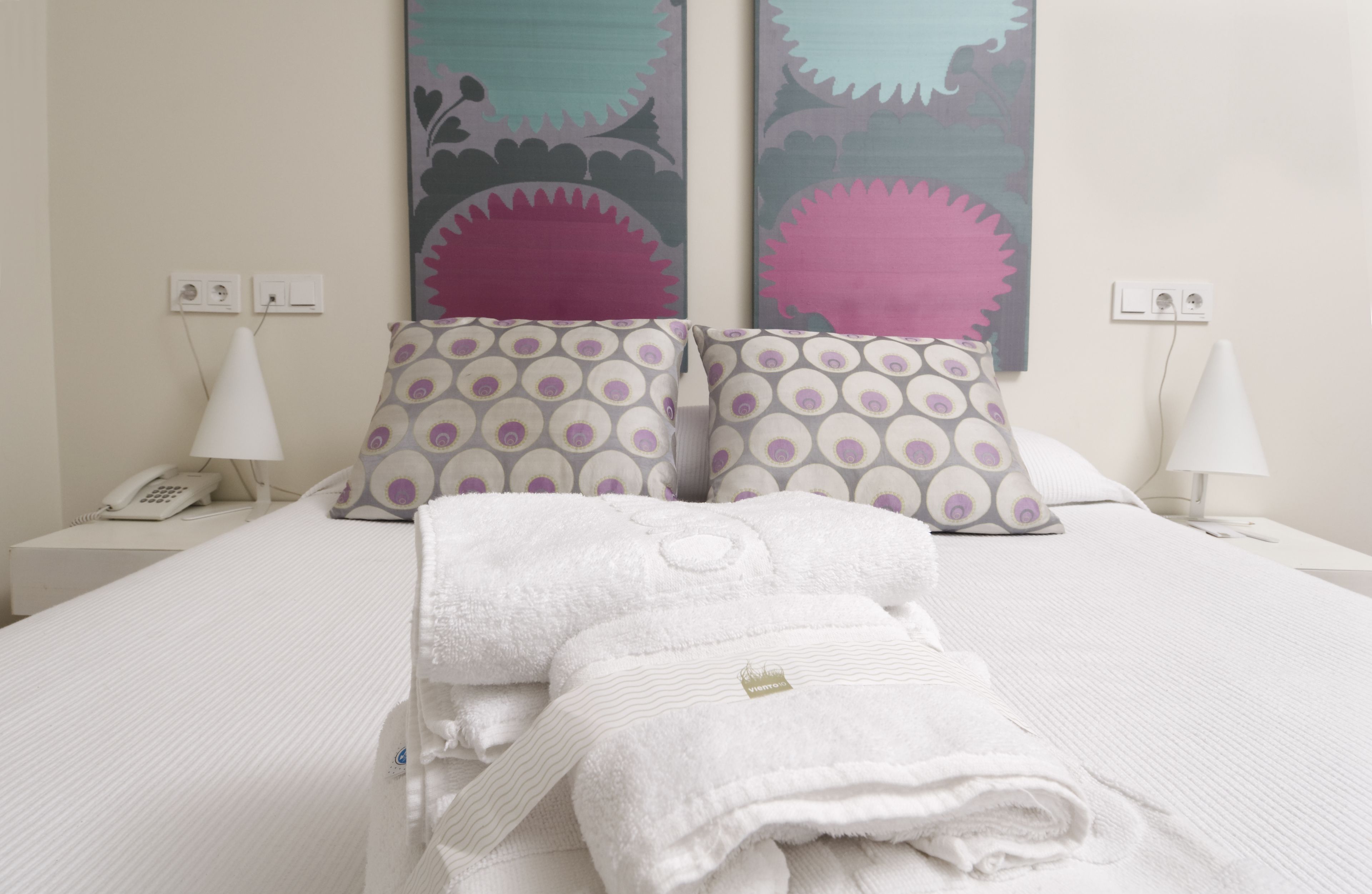 Cama bem arrumada com almofadas estampadas, uma toalha dobrada e um telefone de mesa em um criado-mudo branco.