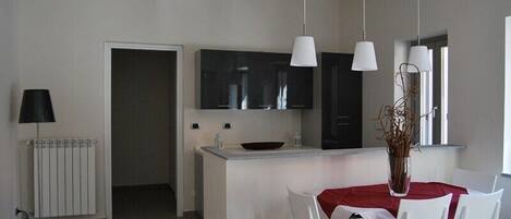Uma cozinha moderna com armários brancos, uma ilha central, luminárias pendentes e uma área de refeições com cadeiras brancas e toalha de mesa vermelha.