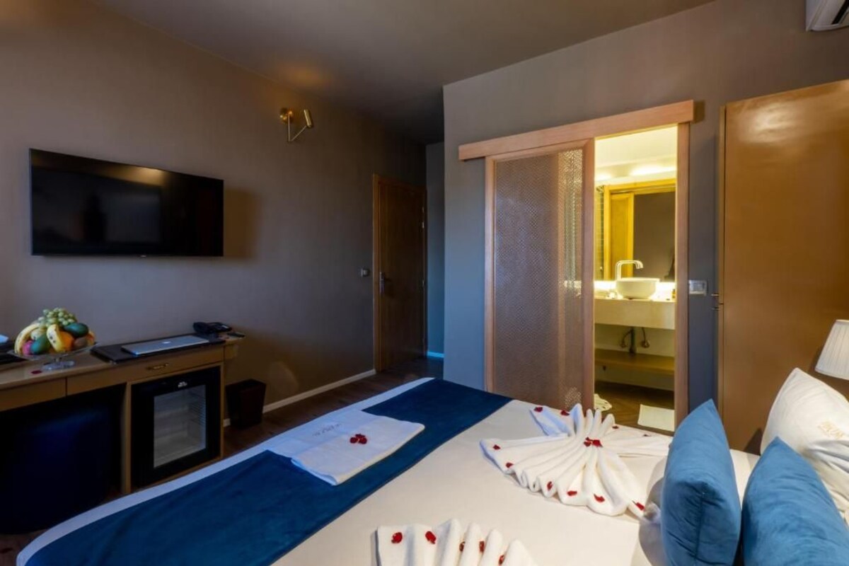 Quarto de hotel com cama, escrivaninha com computador, televisão e banheiro visível por uma porta aberta.