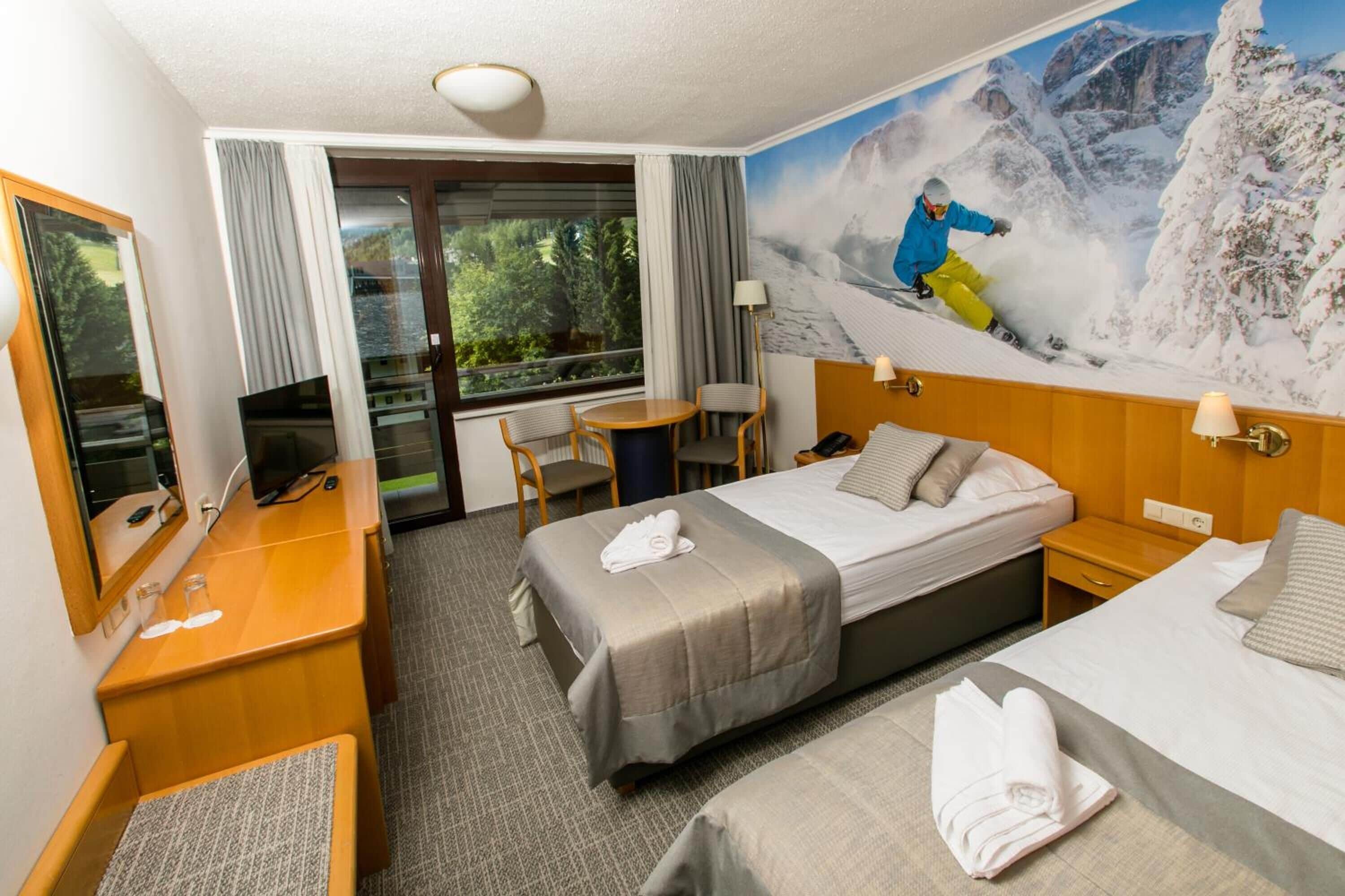 Quarto de hotel com duas camas, uma escrivaninha, uma cadeira, televisão e uma janela grande com vista para as montanhas.