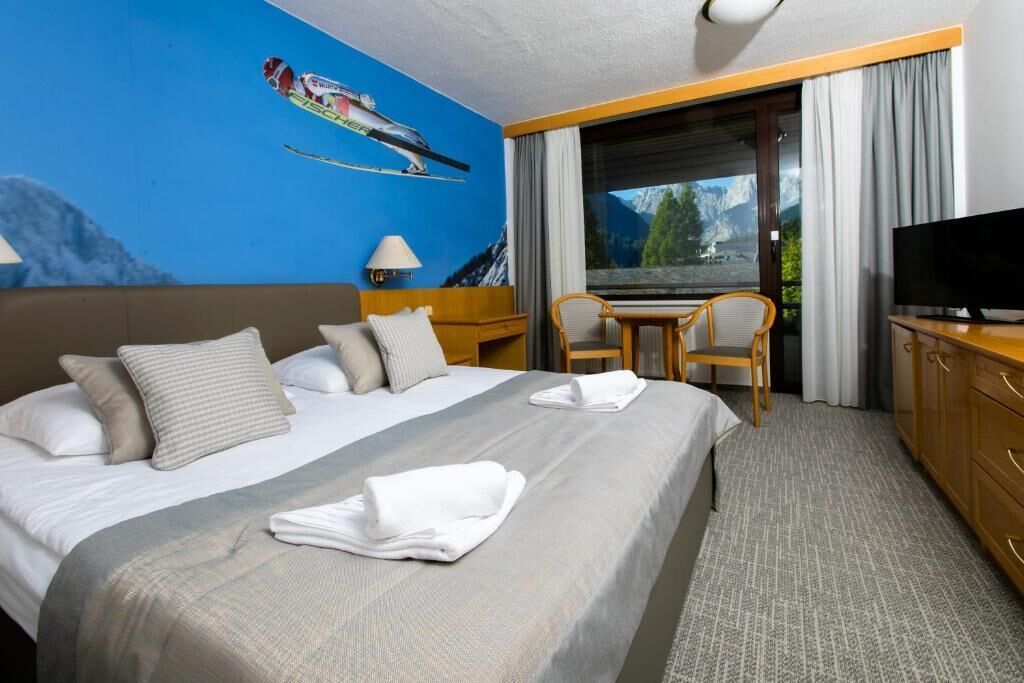 Quarto de hotel com cama, escrivaninha, cadeira, televisão e janela com vista para as montanhas.