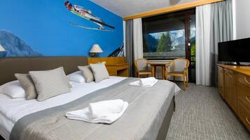 Quarto de hotel com cama, escrivaninha, cadeira, televisão e janela com vista para as montanhas.