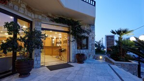 Entrada de hotel com fachada de pedra, grandes vasos de plantas e interior bem iluminado visível pelas portas de vidro.