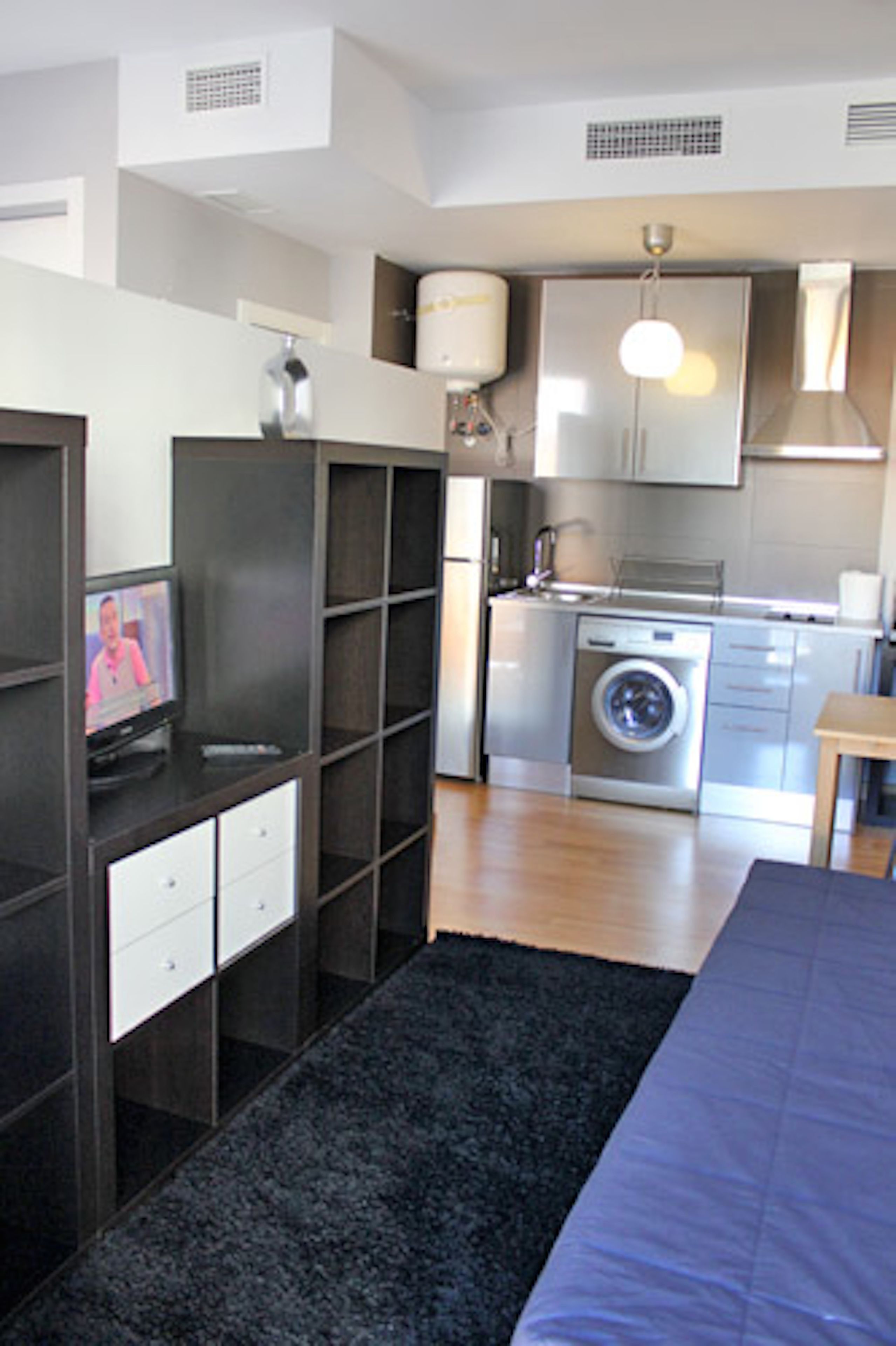 Quarto moderno com uma cama grande, carpete escuro, TV sobre um suporte de madeira, máquina de lavar e área de cozinha com pia e armários.