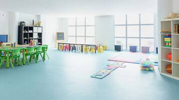 Uma sala de aula luminosa, com cadeiras verdes, uma estante preta e uma janela grande.