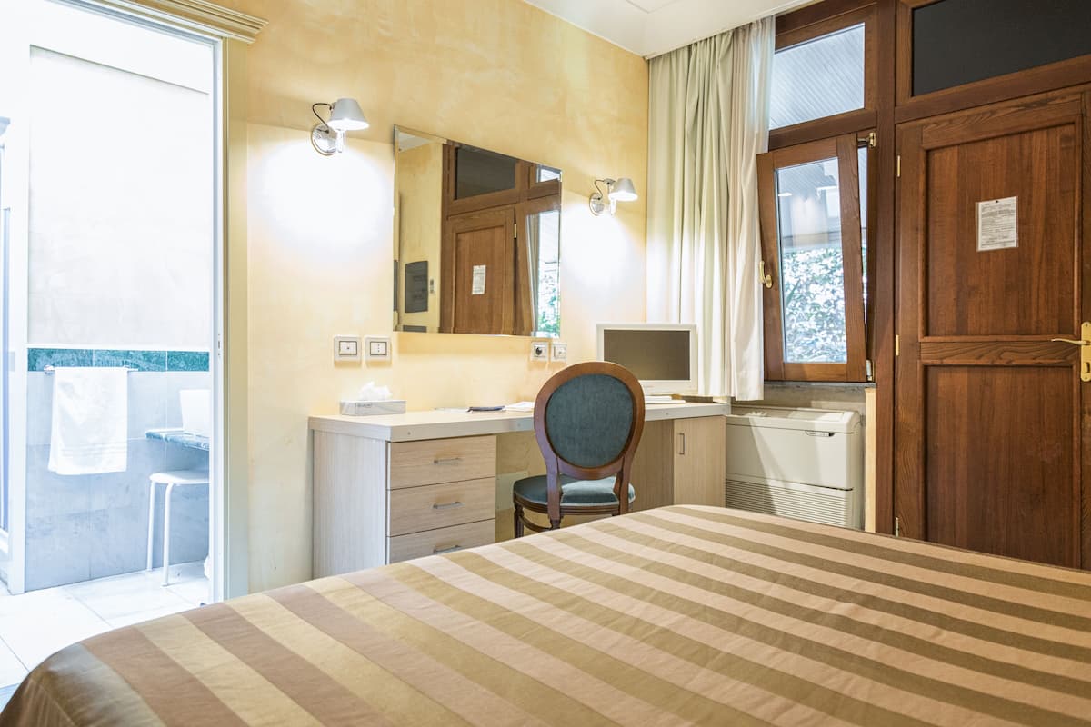 Quarto de hotel com cama, escrivaninha, cadeira, espelho e janela.