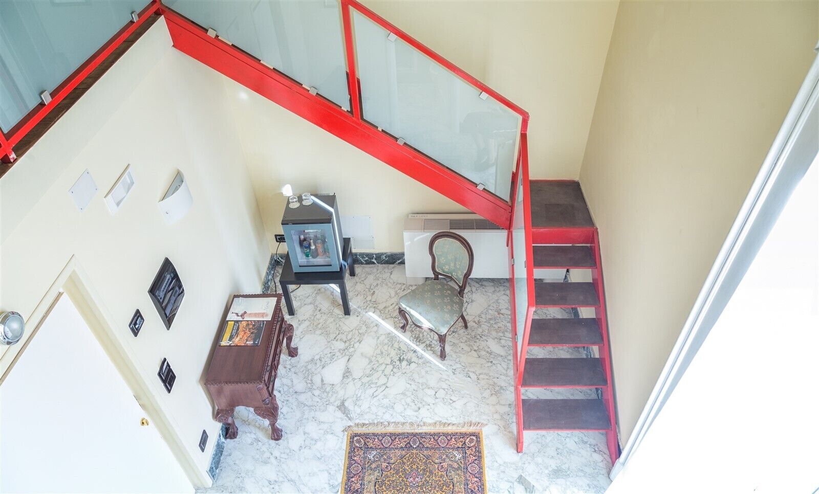 Uma escadaria com corrimão vermelho que leva a um patamar. Há uma cadeira e uma mesa pequena com um abajur no patamar.
