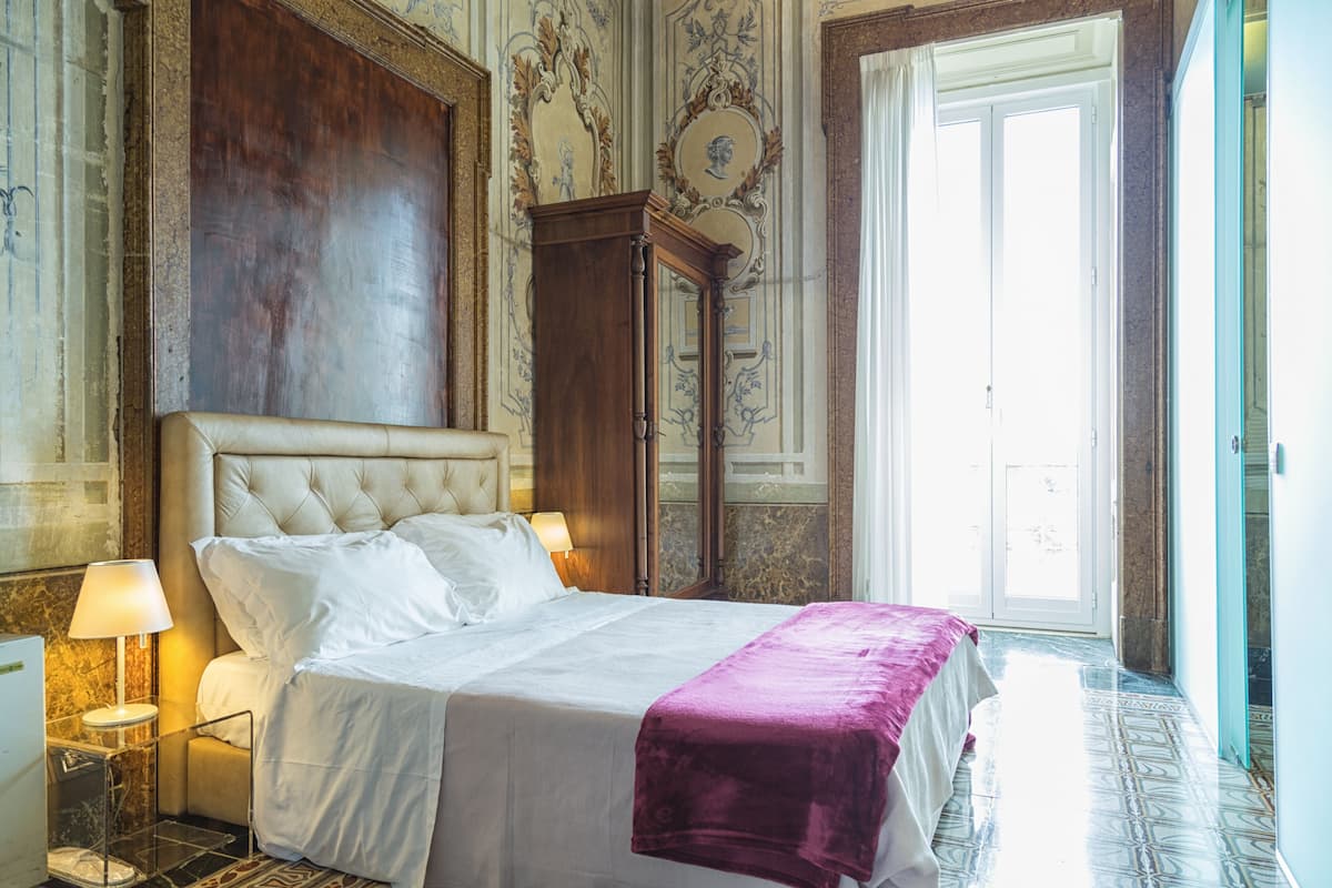 Quarto de hotel com uma cama grande, uma mesa de cabeceira com luminária, um guarda-roupa de madeira e uma janela ampla com vista para o exterior.