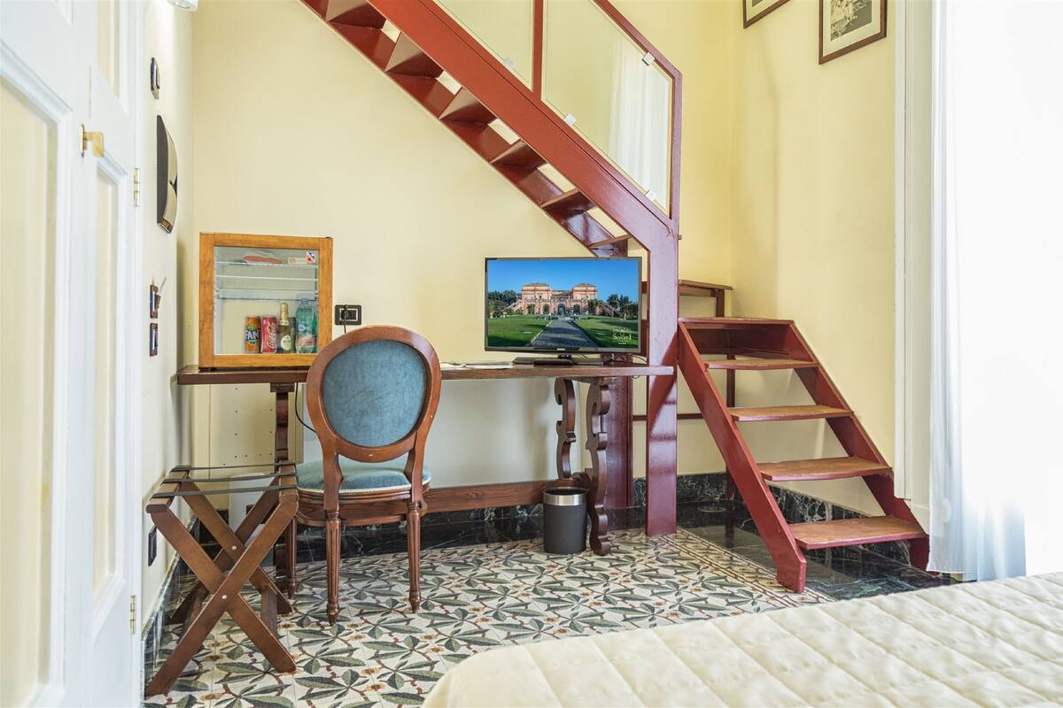 Quarto com cama, escrivaninha com cadeira, televisão, escada vermelha e piso estampado.