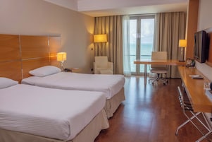 Quarto de hotel com duas camas, uma escrivaninha, uma cadeira, uma TV e vista para o mar.