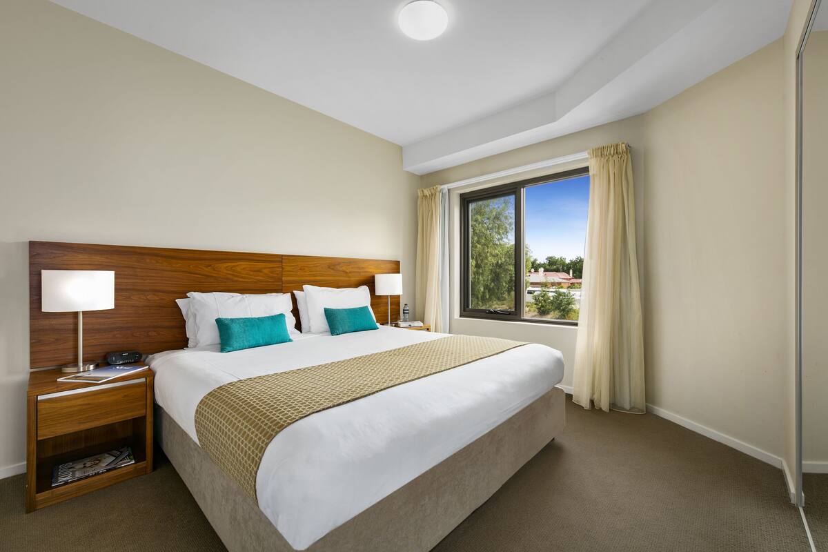 Quarto de hotel com uma cama grande, cabeceira de madeira, duas mesas de cabeceira com abajures, uma janela com cortinas e vista para o verde lá fora.