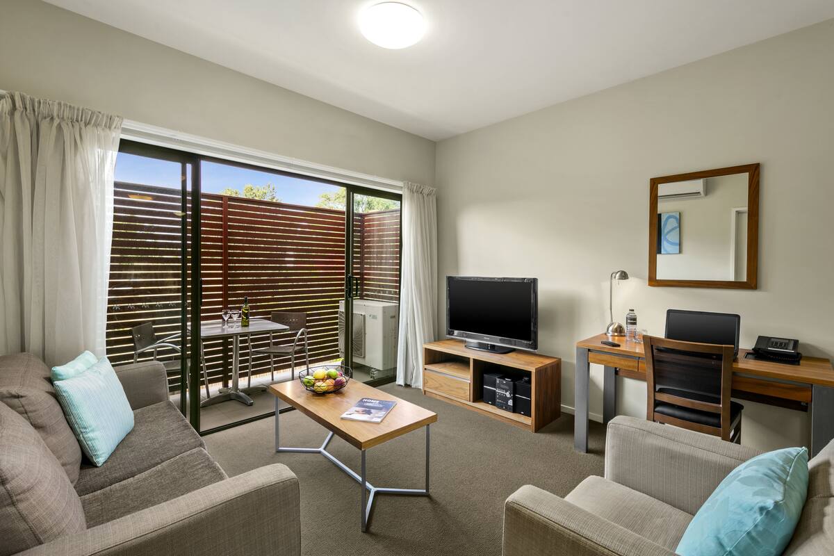 Quarto de hotel moderno com sofá, poltrona, televisão, escrivaninha e área de jantar com mesa e cadeiras.