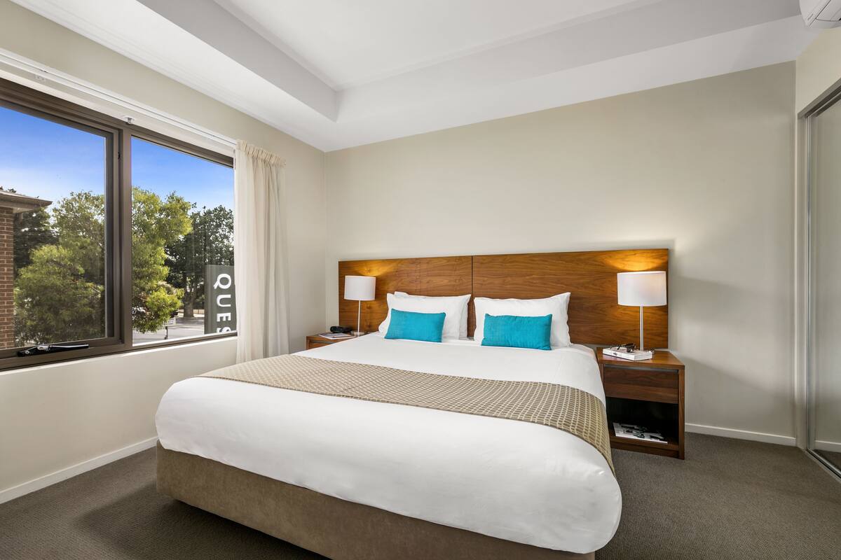 Quarto de hotel com uma cama grande, duas mesas de cabeceira, uma janela com cortinas e vista para árvores e um edifício de tijolos.
