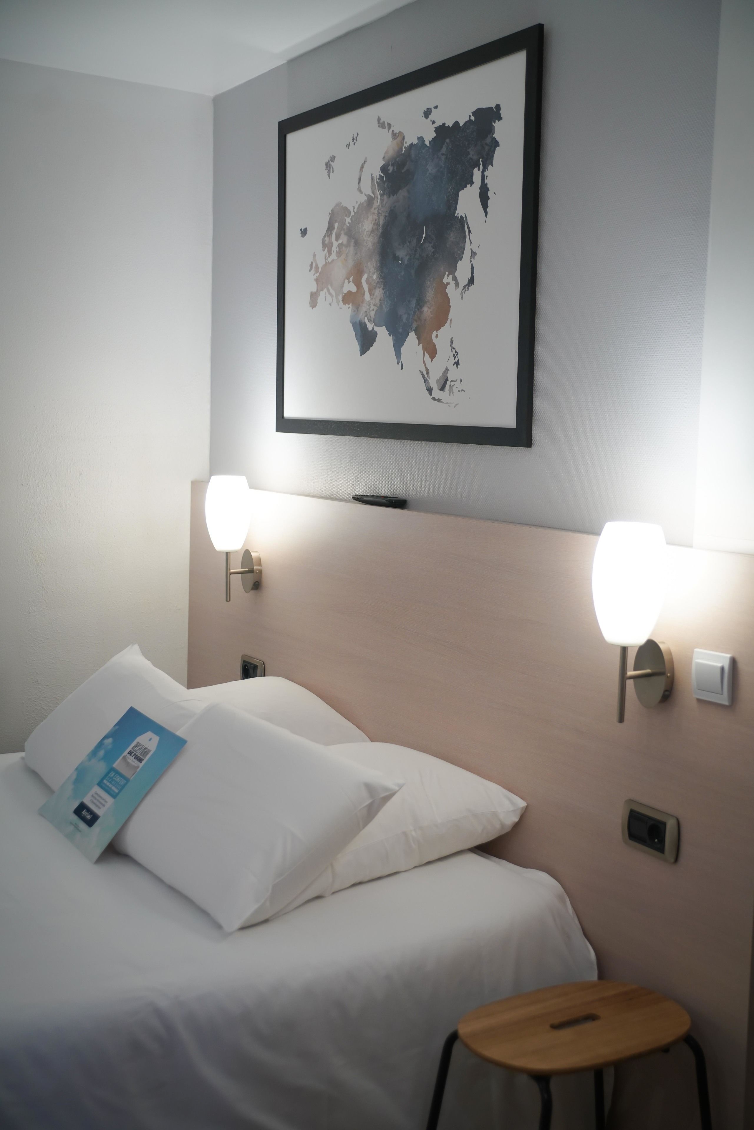 Cama bem arrumada com lençóis brancos, um pequeno banco de madeira e duas luminárias de leitura fixadas na parede.