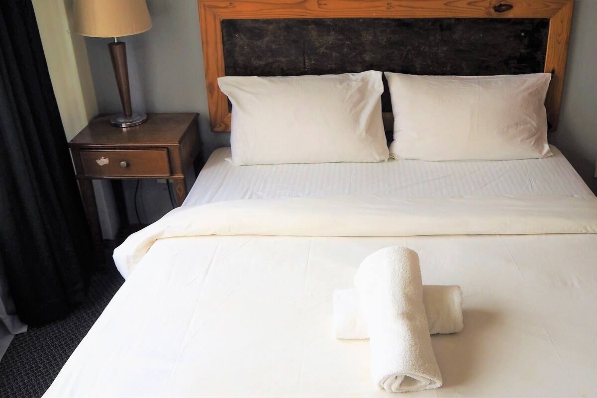 Cama bem arrumada com lençóis e almofadas brancas, mesa de cabeceira de madeira com abajur e uma tomada na parede.