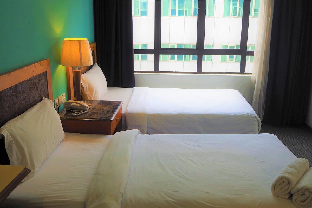 Quarto de hotel com duas camas, uma mesa de cabeceira com abajur e uma janela com cortinas.
