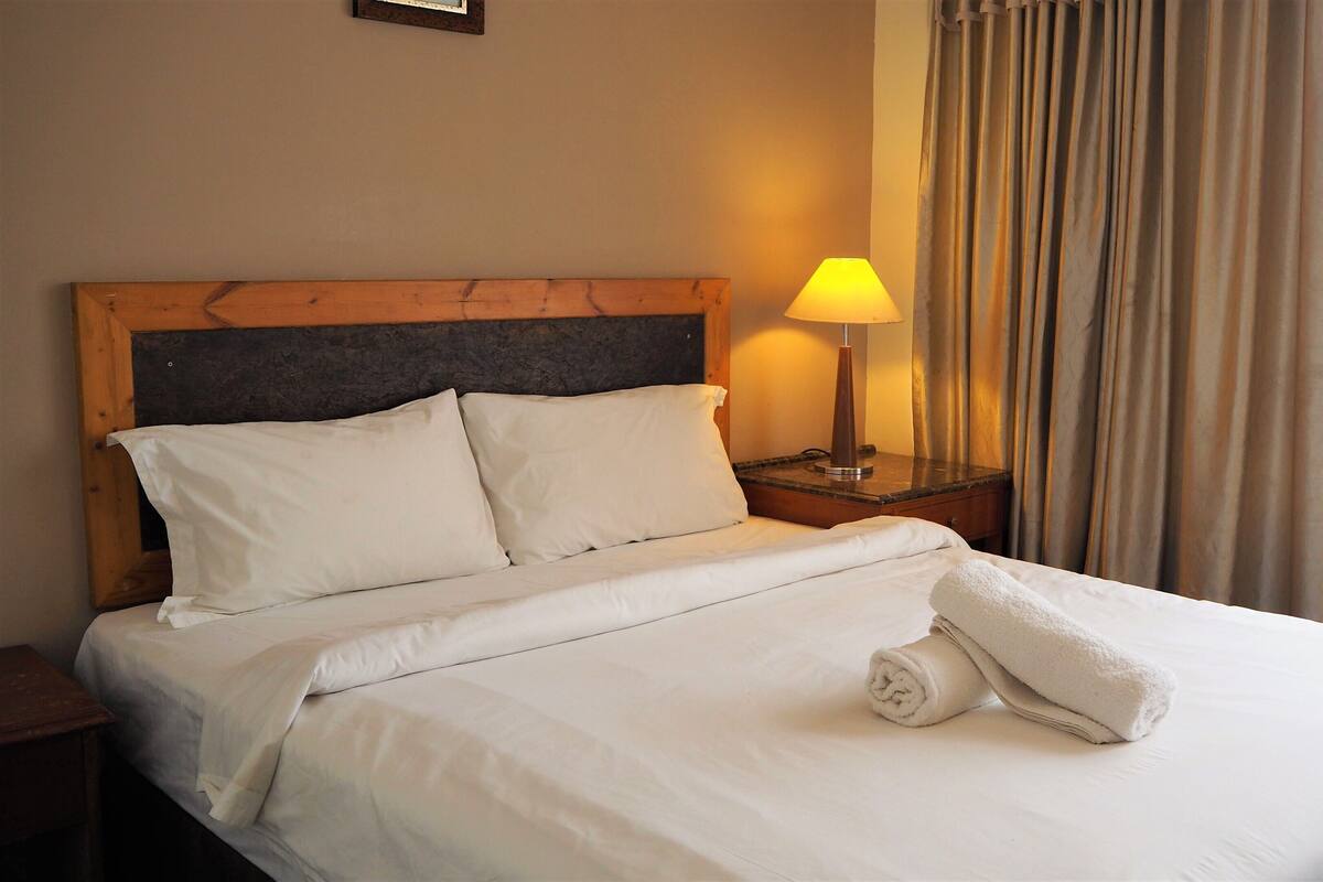 Uma cama bem arrumada com lençóis brancos, cabeceira de madeira, abajur e toalha enrolada sobre a cama.
