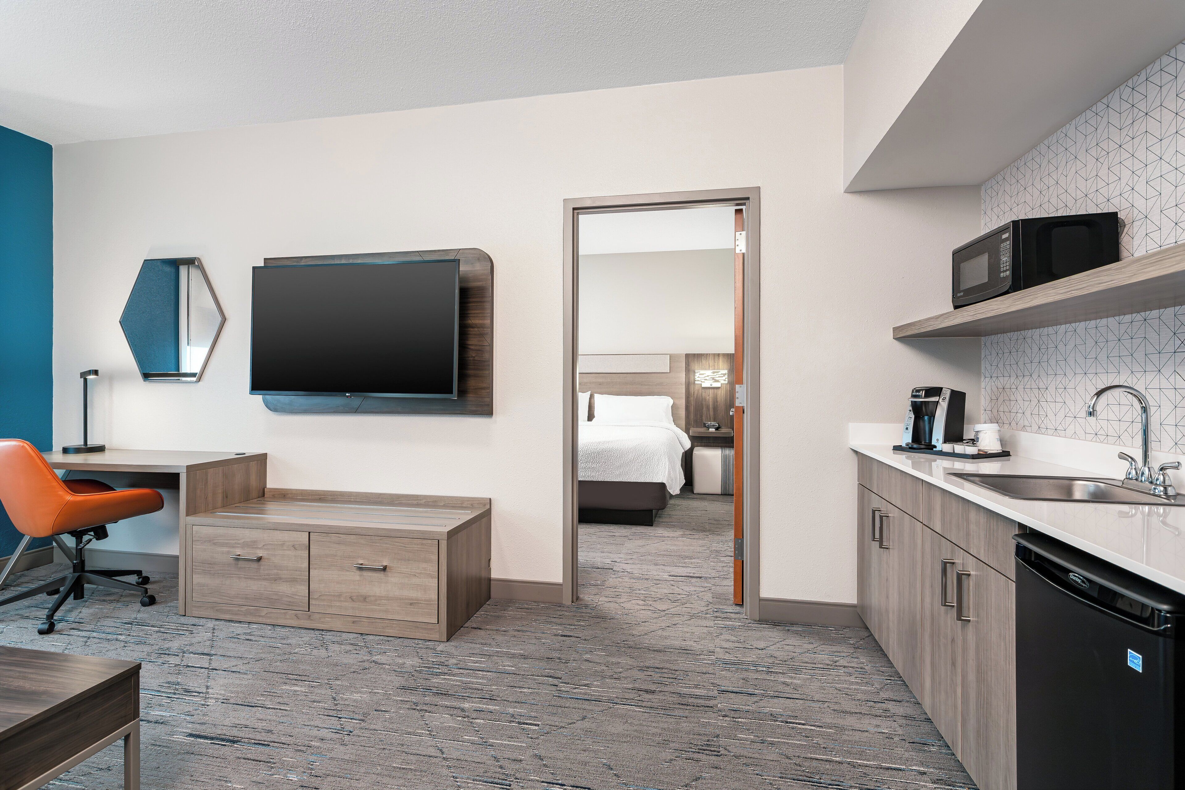Quarto de hotel moderno com cozinha compacta, televisão de tela plana, escrivaninha com cadeira e cama com edredom branco.