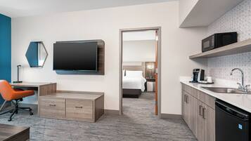 Quarto de hotel moderno com cozinha compacta, televisão de tela plana, escrivaninha com cadeira e cama com edredom branco.