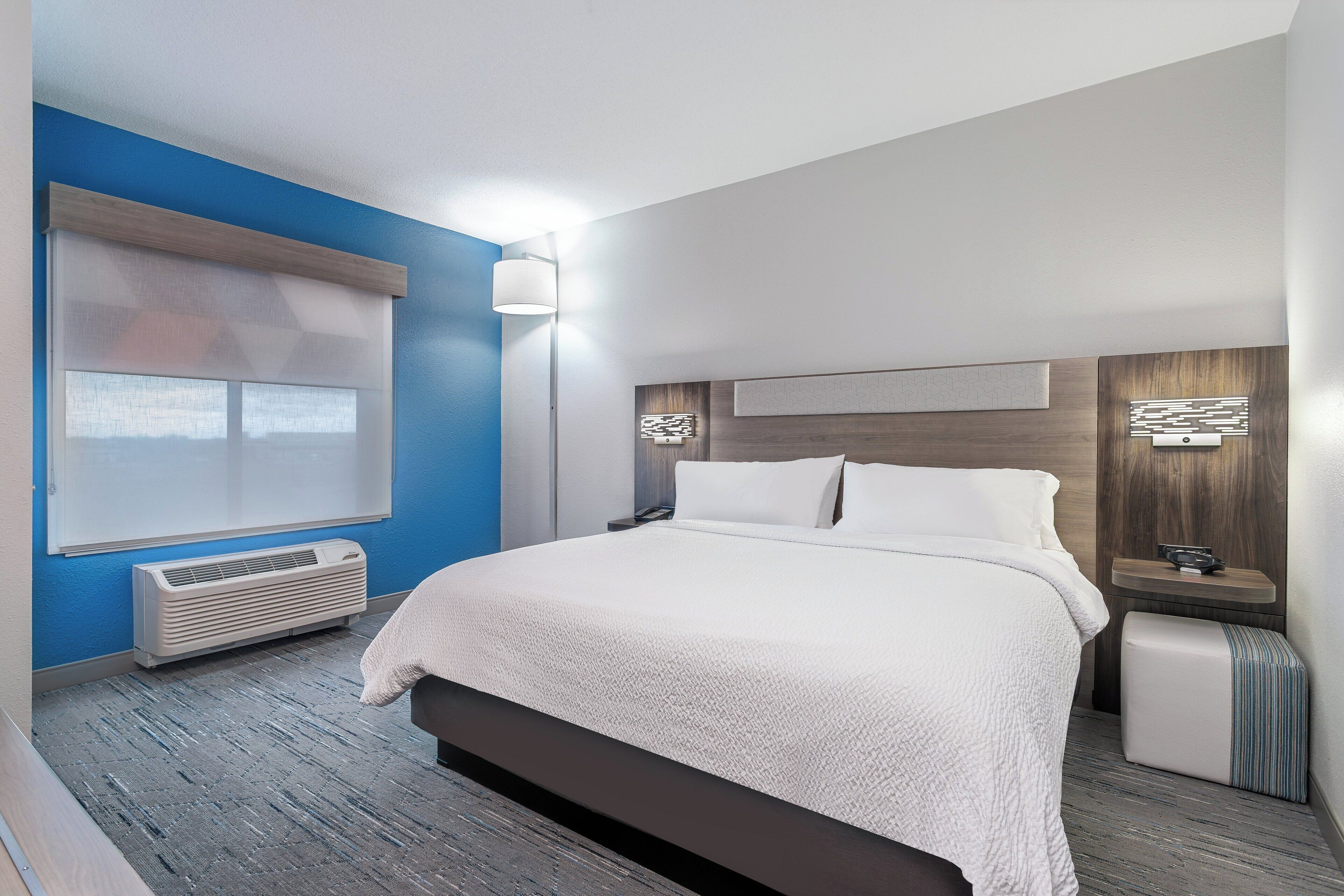 Quarto de hotel moderno com cama grande, janela com persianas, luminária de cabeceira e mesa de cabeceira pequena.