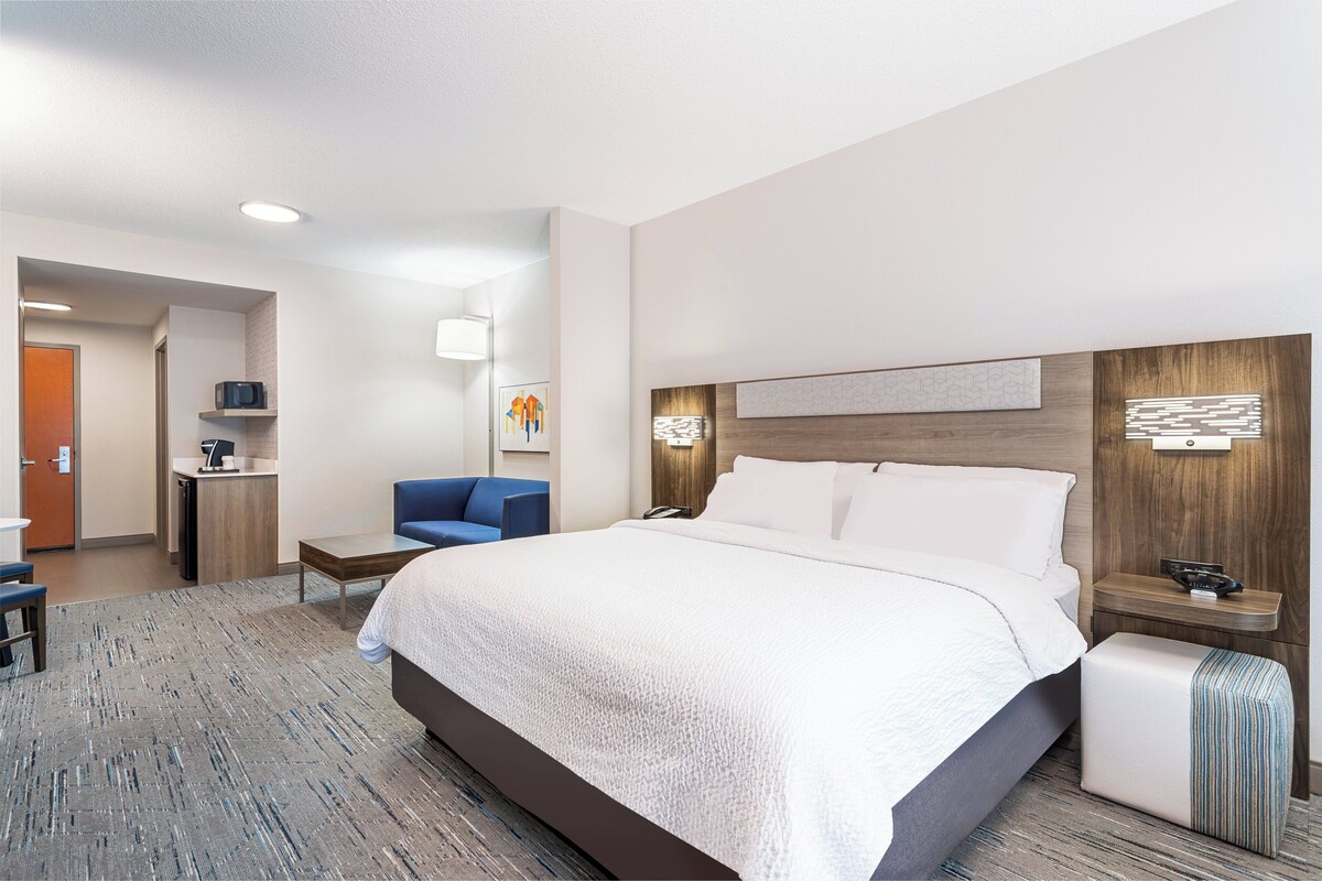 Quarto de hotel com uma cama grande, uma escrivaninha com cadeira, uma kitchenette pequena e um banheiro.