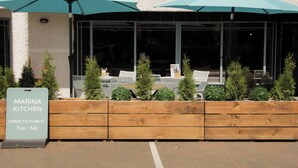 Um jardim em vasos de madeira com vegetação, área de estar externa com guarda-sóis e placa indicando 'Marina Kitchen' aberta ao público.