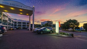 Entrada de hotel com uma placa indicando "ANCHORAGE", área de estacionamento com vários carros e céu claro com cores do pôr do sol.