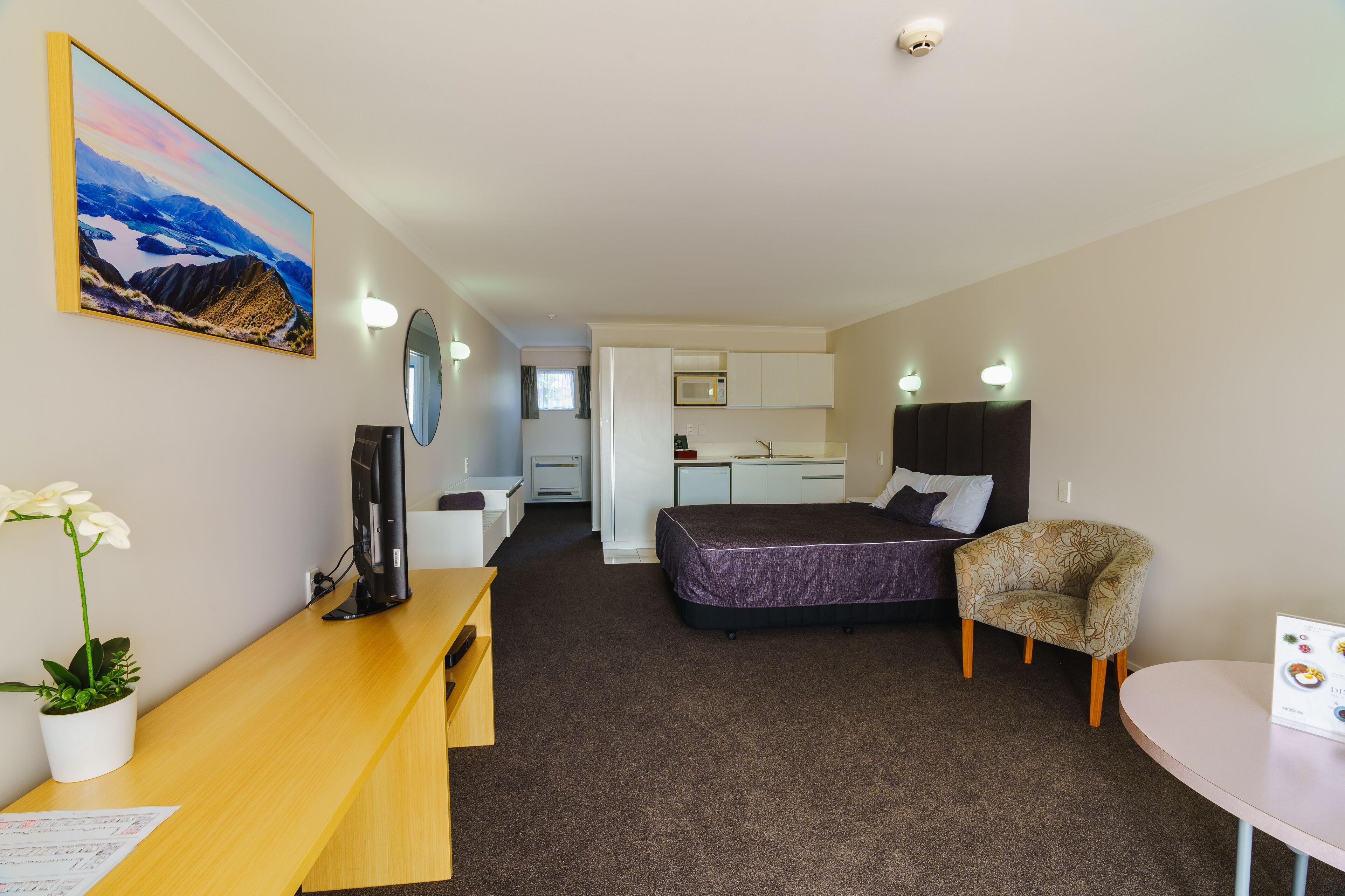 Quarto de hotel com cama, escrivaninha, cadeira e cozinha compacta.