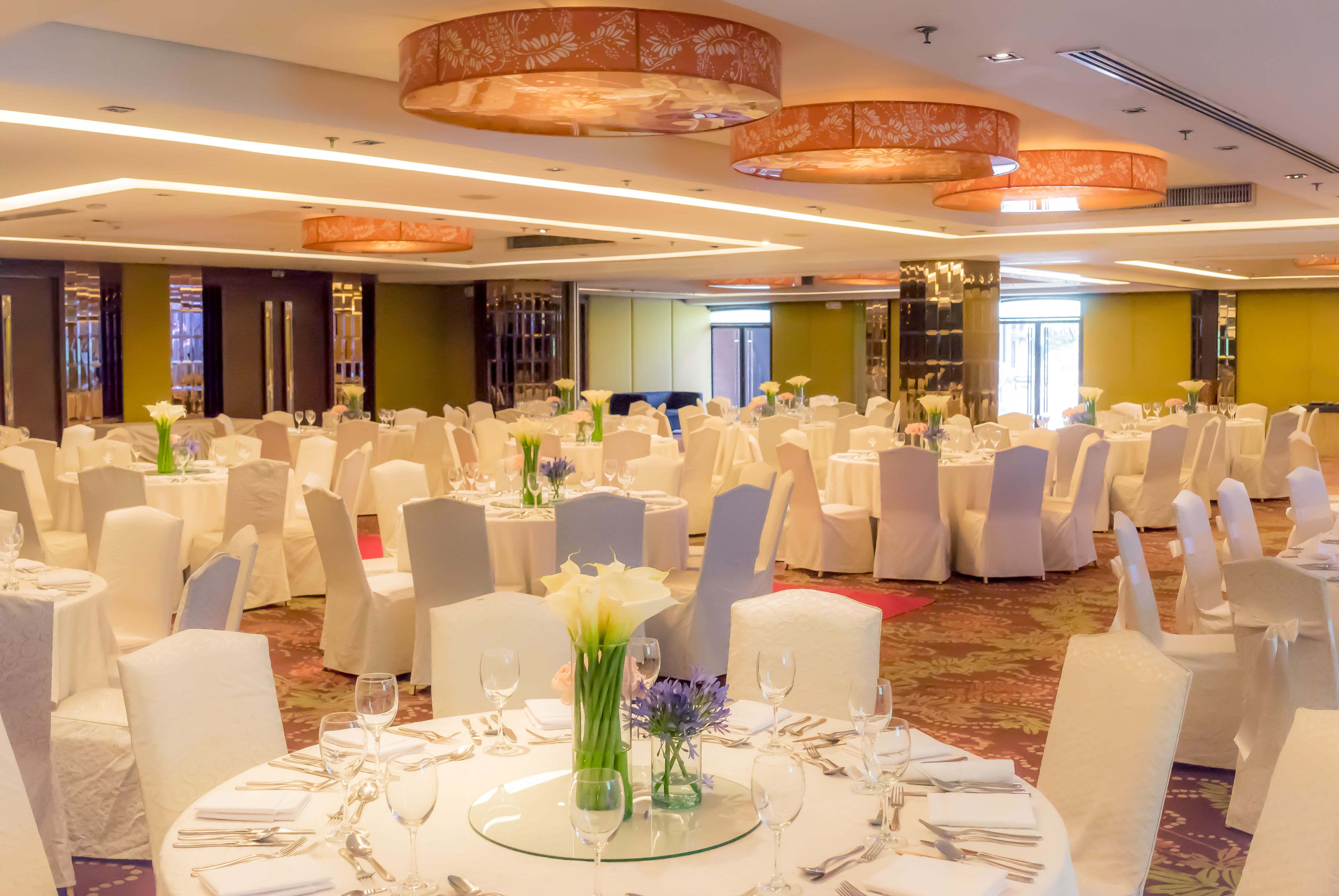 Salão de banquetes com mesas redondas postas para refeições, cadeiras brancas e centros de mesa florais.