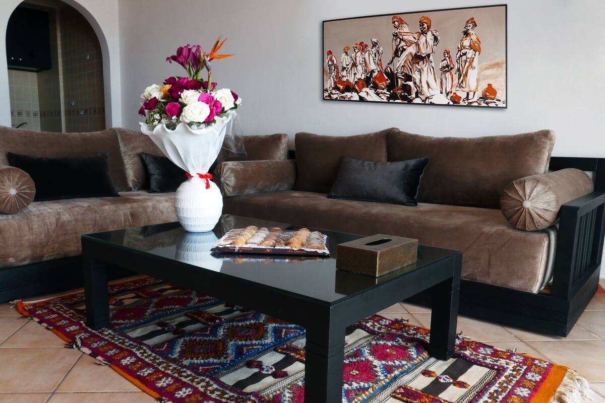 Uma sala de estar com um sofá, uma mesa de centro, um vaso com flores, um quadro emoldurado e um tapete estampado.