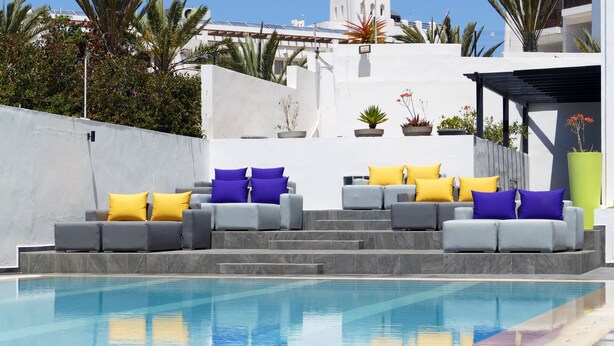 Uma área à beira da piscina com cadeiras de lounge cinzentas e almofadas amarelas e roxas, cercada por um muro branco e plantas em vasos.