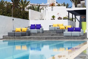 Uma área à beira da piscina com cadeiras de lounge cinzentas e almofadas amarelas e roxas, cercada por um muro branco e plantas em vasos.