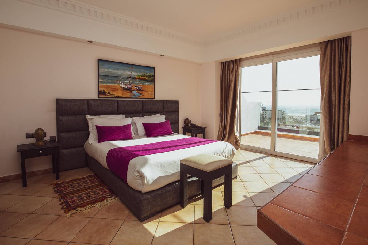 Quarto com cama grande, mesa de cabeceira, cortinas na janela e um quadro na parede.