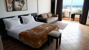 Quarto de hotel com uma cama grande, um sofá, uma mesinha e vista para o mar através de uma porta de correr.