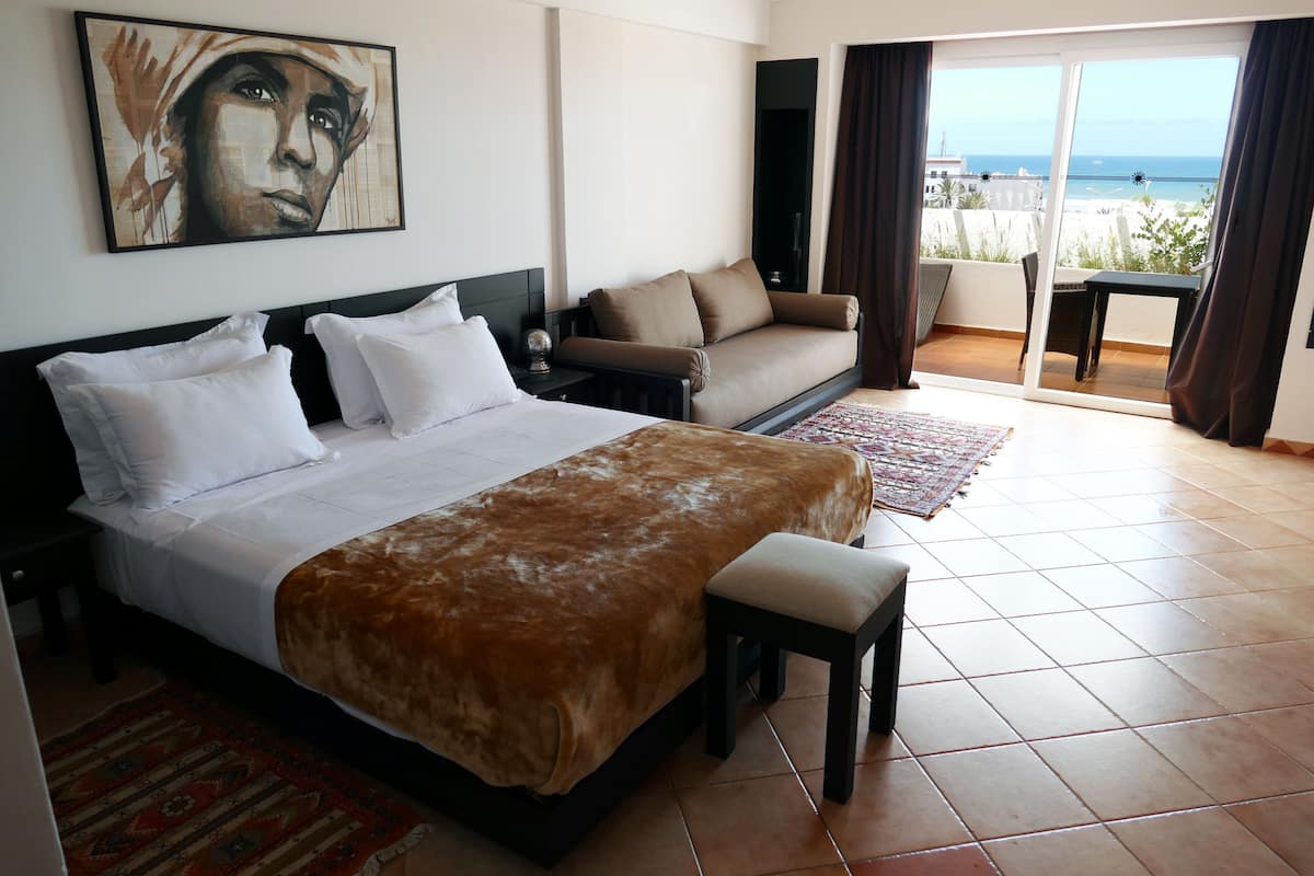 Quarto de hotel com uma cama grande, um sofá, uma mesinha e vista para o mar através de uma porta de correr.