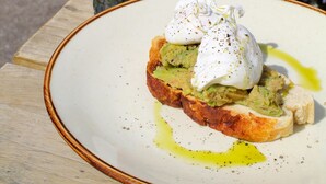 Um prato de comida com ovo escaldado e abacate na torrada, um pequeno vaso com flores e uma mesa de madeira.