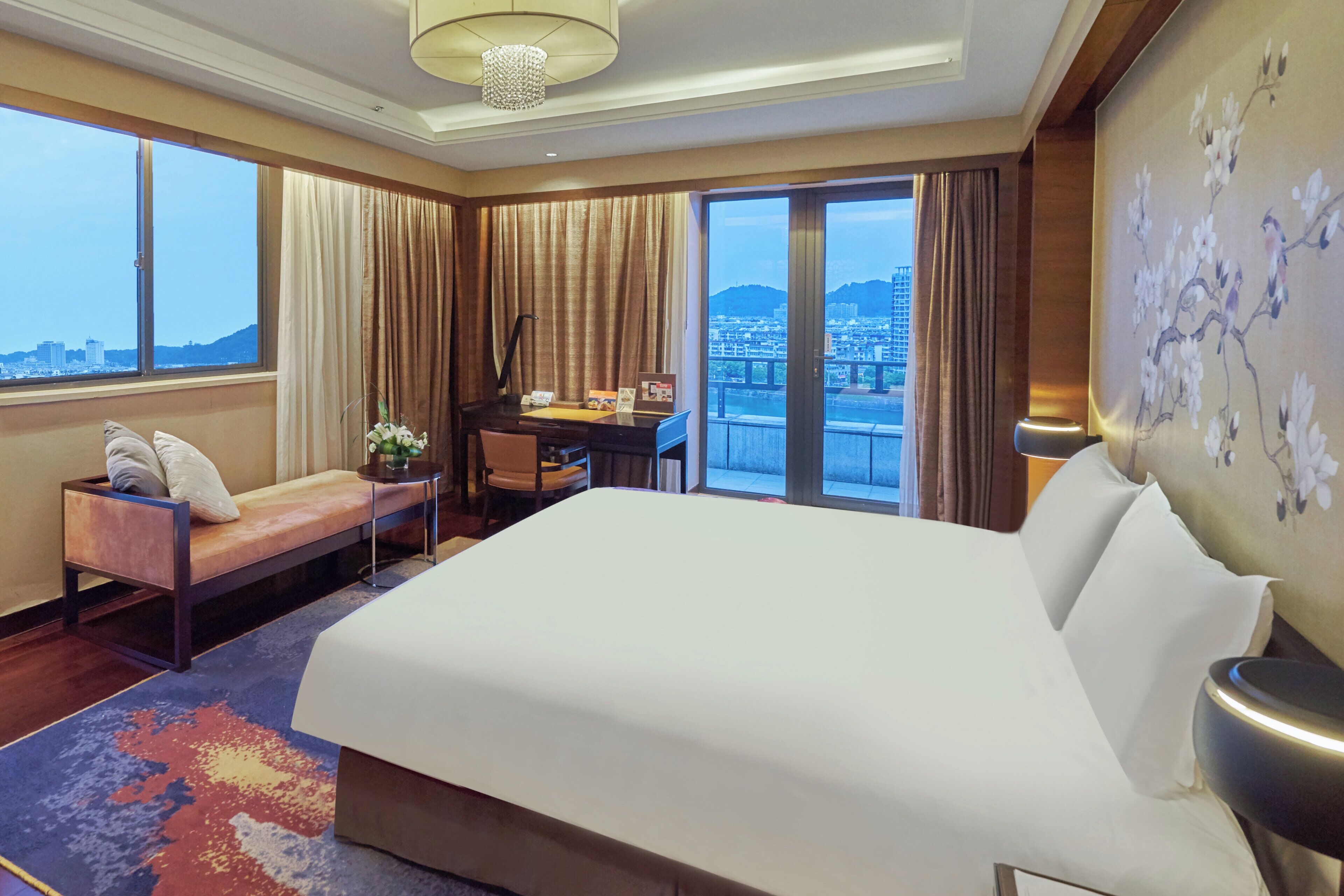 Quarto de hotel com uma cama grande, uma escrivaninha, uma cadeira, um sofá e vista para a cidade através de amplas janelas.