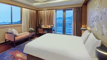 Quarto de hotel com uma cama grande, uma escrivaninha, uma cadeira, um sofá e vista para a cidade através de amplas janelas.