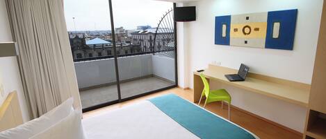 Quarto de hotel com cama, escrivaninha, cadeira, televisão e vista para a cidade através de amplas janelas.
