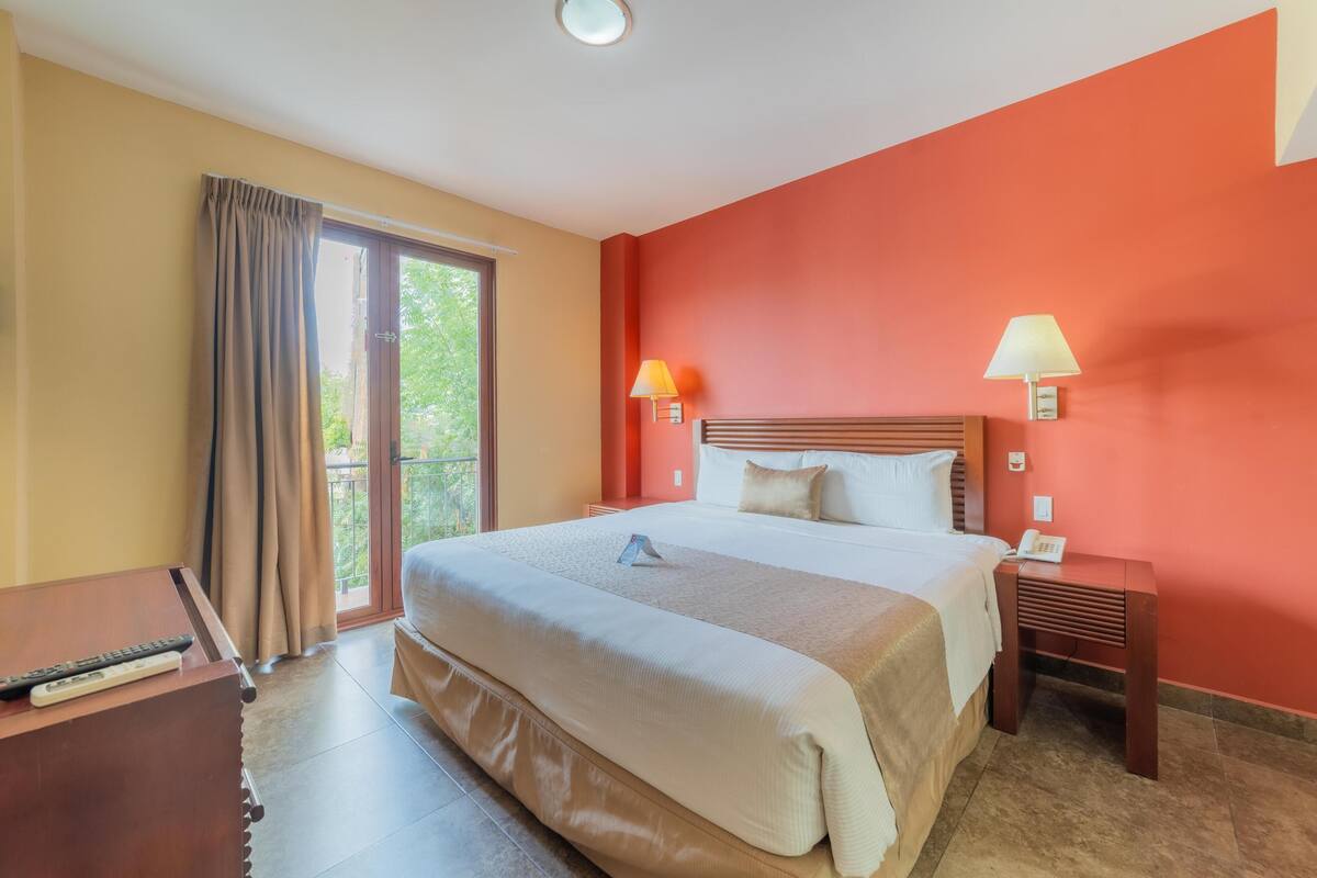 Quarto de hotel com cama, luminárias de cabeceira, escrivaninha e cortinas na janela.