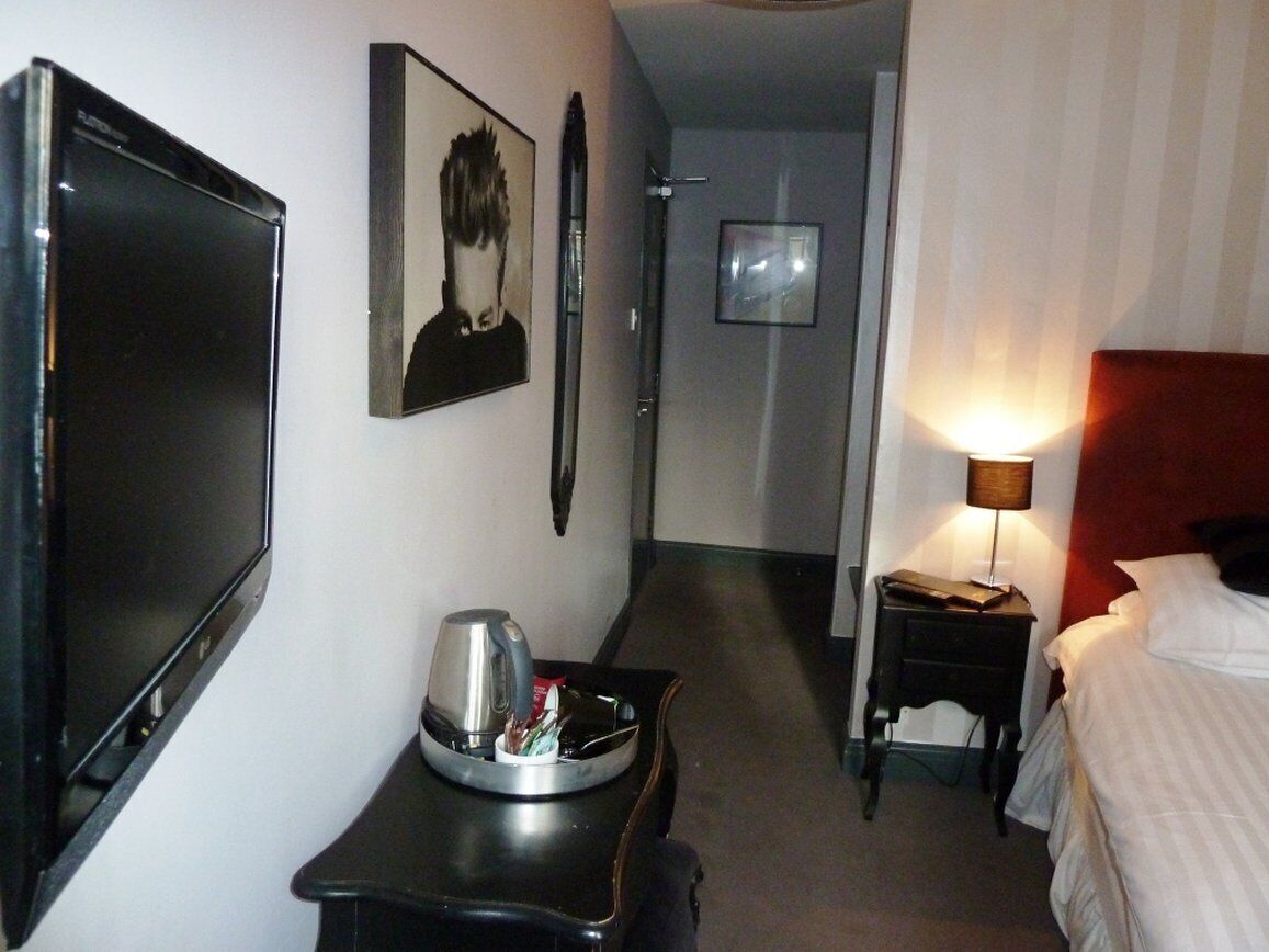 Quarto de hotel com uma cama, uma mesa com luminária, uma televisão e um quadro na parede.