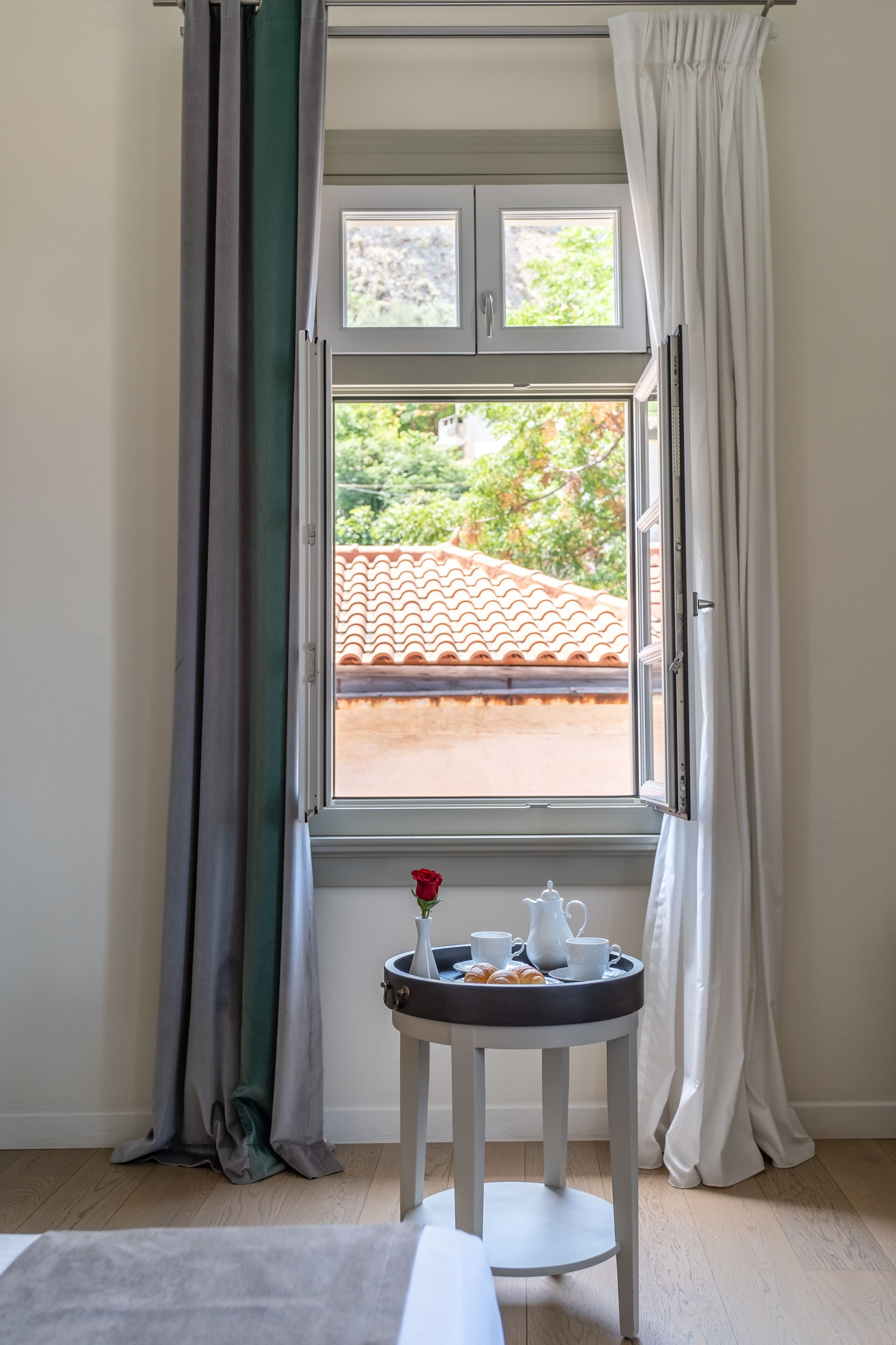 Quarto com janela, uma pequena mesa com um vaso e chaleira, e vista para um telhado de telhas lá fora.