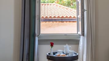 Quarto com janela, uma pequena mesa com um vaso e chaleira, e vista para um telhado de telhas lá fora.
