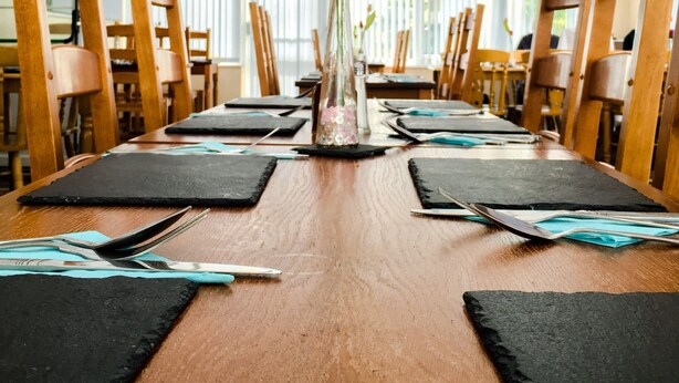 Uma área de refeições com uma mesa de madeira comprida posta para uma refeição, rodeada por cadeiras de madeira.