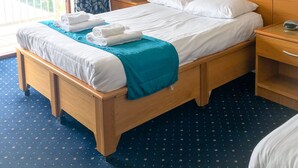 Uma cama bem arrumada com lençóis brancos e um cobertor azul-turquesa, um criado-mudo de madeira com um relógio, uma janela com cortinas azuis e uma pintura de paisagem urbana na parede.