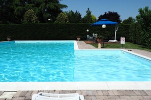 Piscina retangular com água azul límpida, rodeada por um deck pavimentado. Há um guarda-sol azul e uma cadeira branca perto da piscina.