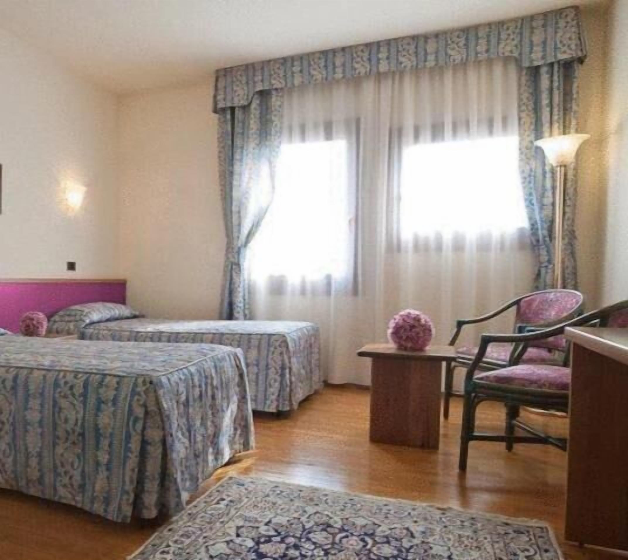 Quarto de hotel com duas camas, uma mesa pequena, uma cadeira e um abajur.