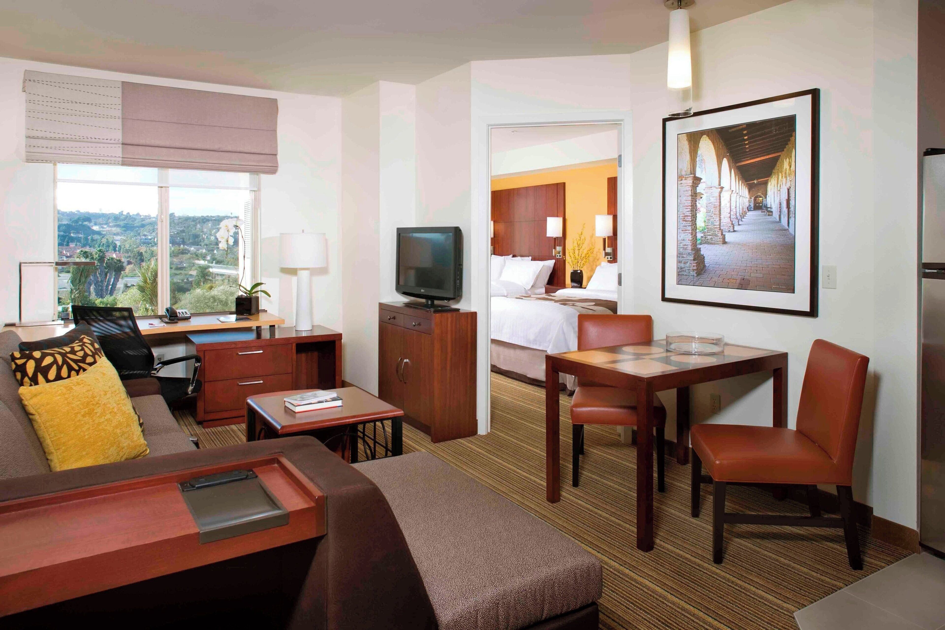 Quarto de hotel com sofá, escrivaninha com computador, televisão, mesa de jantar com cadeiras e cama.