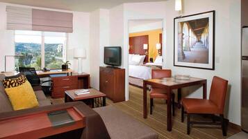 Quarto de hotel com sofá, escrivaninha com computador, televisão, mesa de jantar com cadeiras e cama.